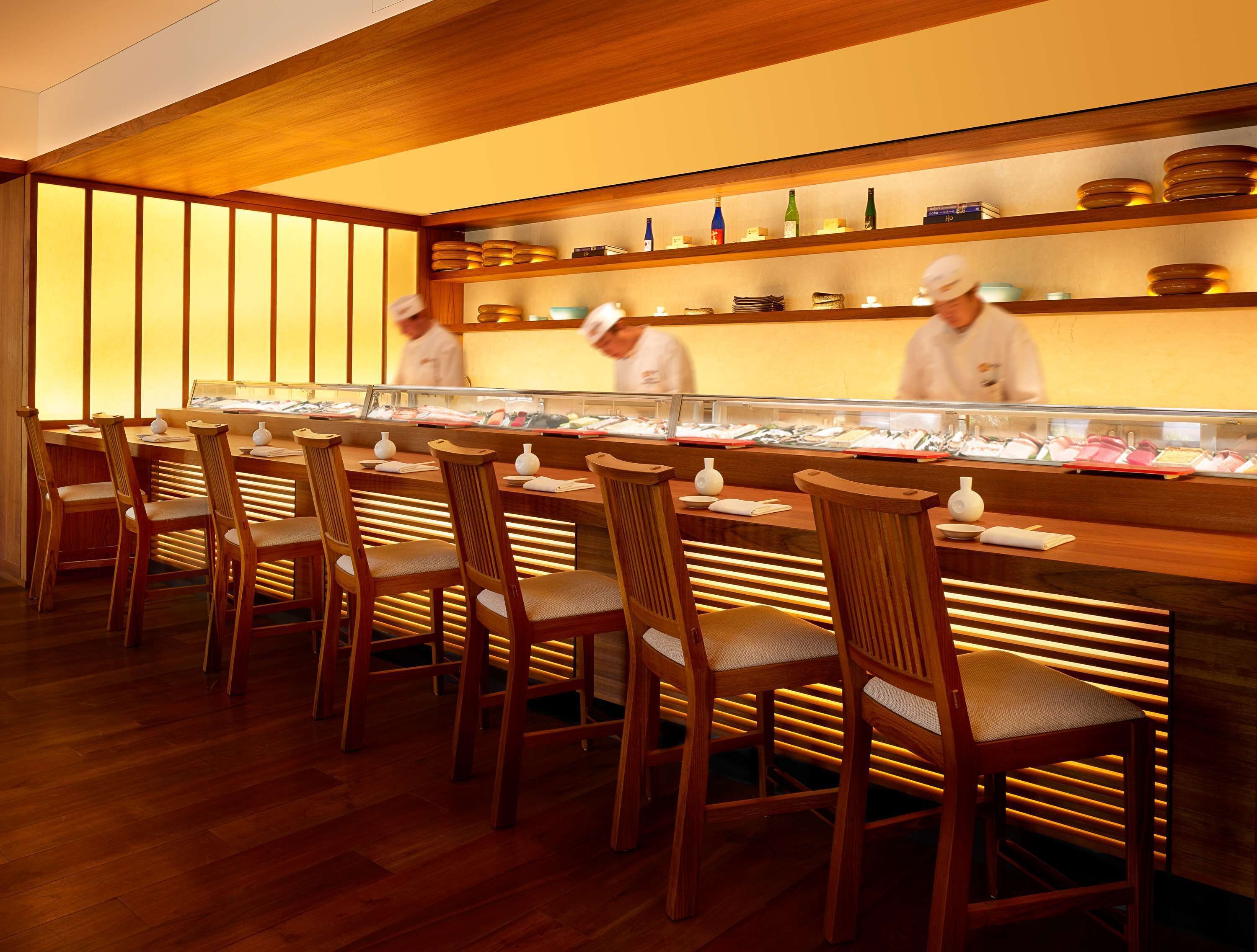Um restaurante com cadeiras e mesas de madeira, um balcão de sushi e uma área de preparo de alimentos.