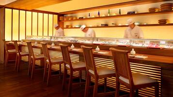 Um restaurante com cadeiras e mesas de madeira, um balcão de sushi e uma área de preparo de alimentos.