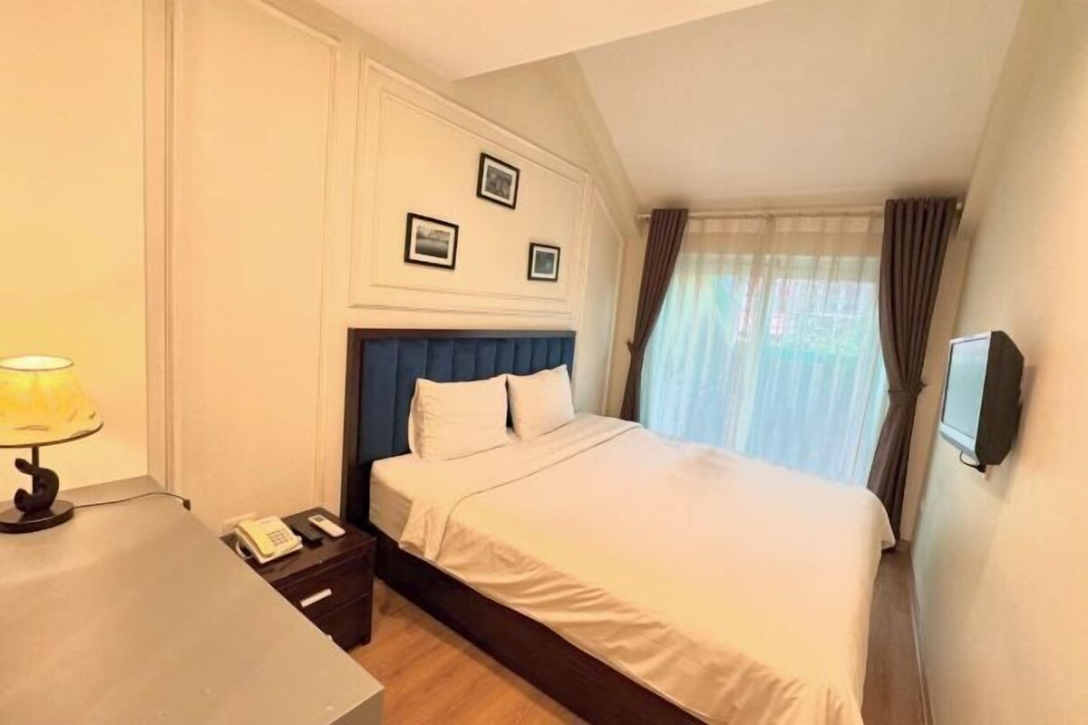Quarto de hotel com cama, mesa de cabeceira, luminária, telefone e televisão na parede.