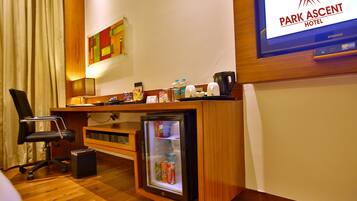 Bancada de recepção de hotel com um refrigerador contendo bebidas, uma televisão exibindo o nome do hotel e uma mesa de madeira com diversos objetos.