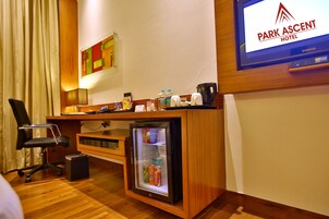 Bancada de recepção de hotel com um refrigerador contendo bebidas, uma televisão exibindo o nome do hotel e uma mesa de madeira com diversos objetos.
