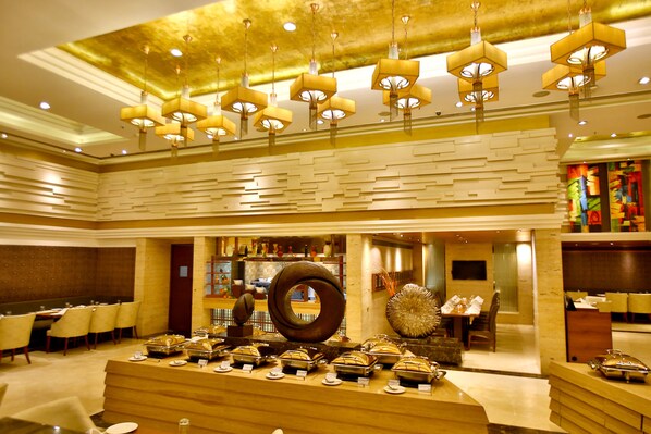 Um saguão de hotel com uma área de buffet central, diversos arranjos de assento e um design moderno de lustre.