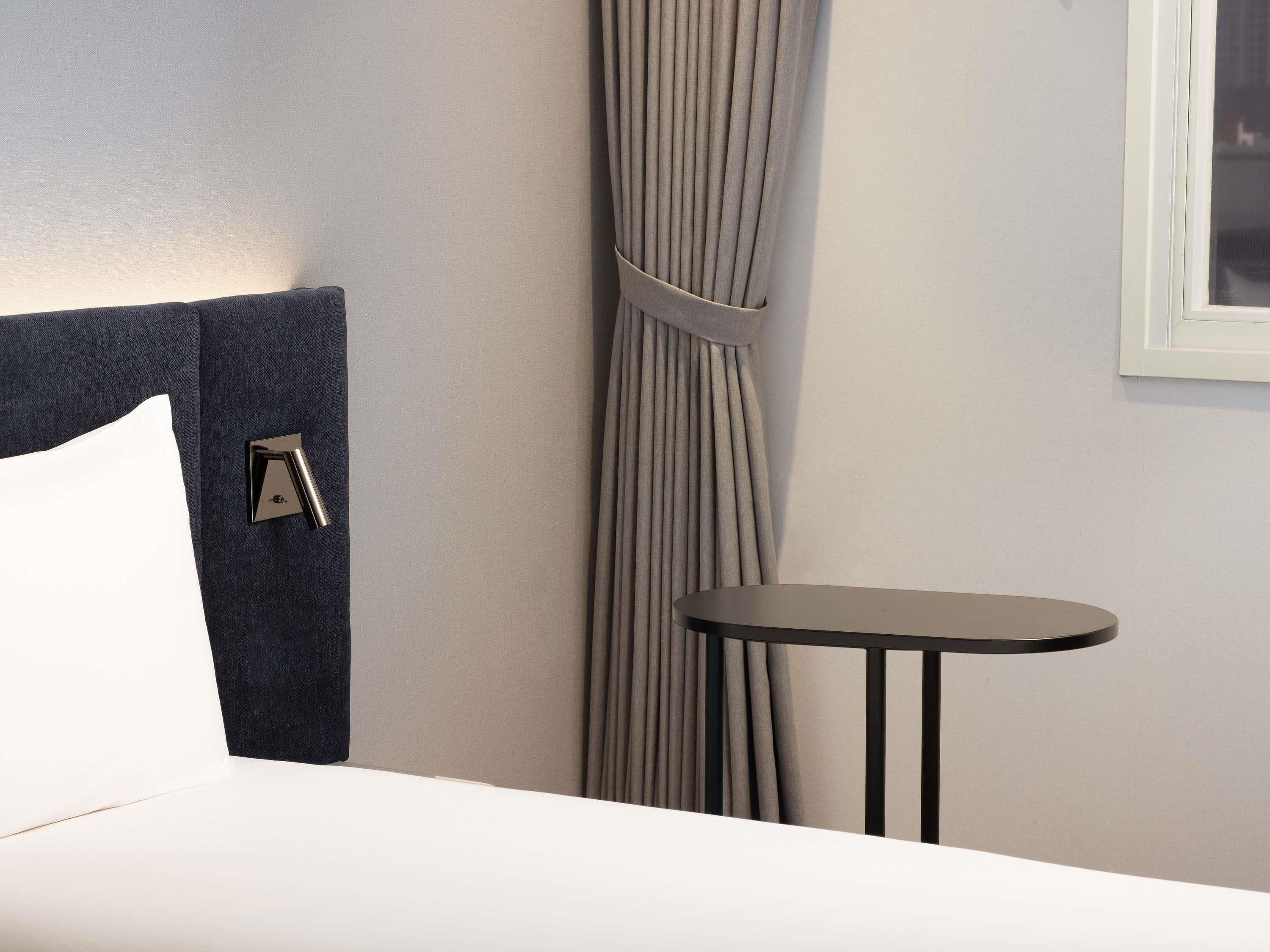 Quarto de hotel com cama, uma pequena mesa, cortina e luminária fixada na parede.