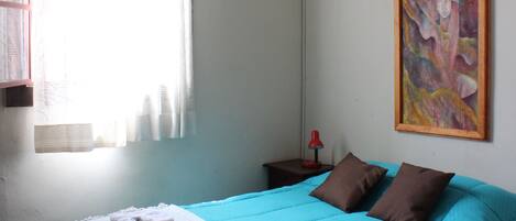 Quarto com cama, mesa de cabeceira, luminária, quadro e janela com cortinas.