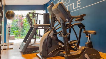 Ginásio com equipamentos de exercício, incluindo esteira e bicicleta ergométrica, parede azul e vista para o exterior arborizado.