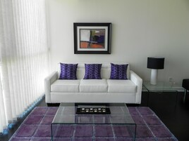 Um sofá branco com três almofadas roxas estampadas, uma mesa de centro de vidro com objetos decorativos, um quadro abstrato emoldurado e um abajur sobre uma mesinha lateral.