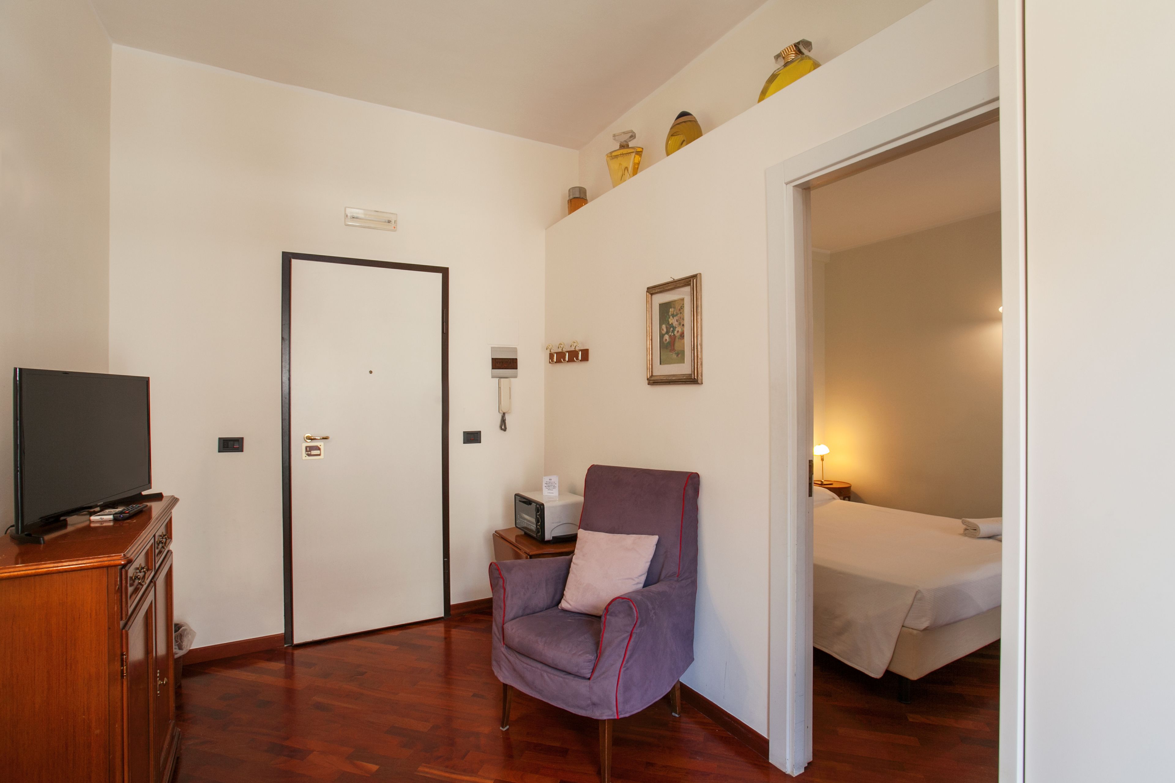 Quarto de hotel com piso de madeira, televisão em um móvel, uma cadeira, mesa de cabeceira com abajur e uma cama.