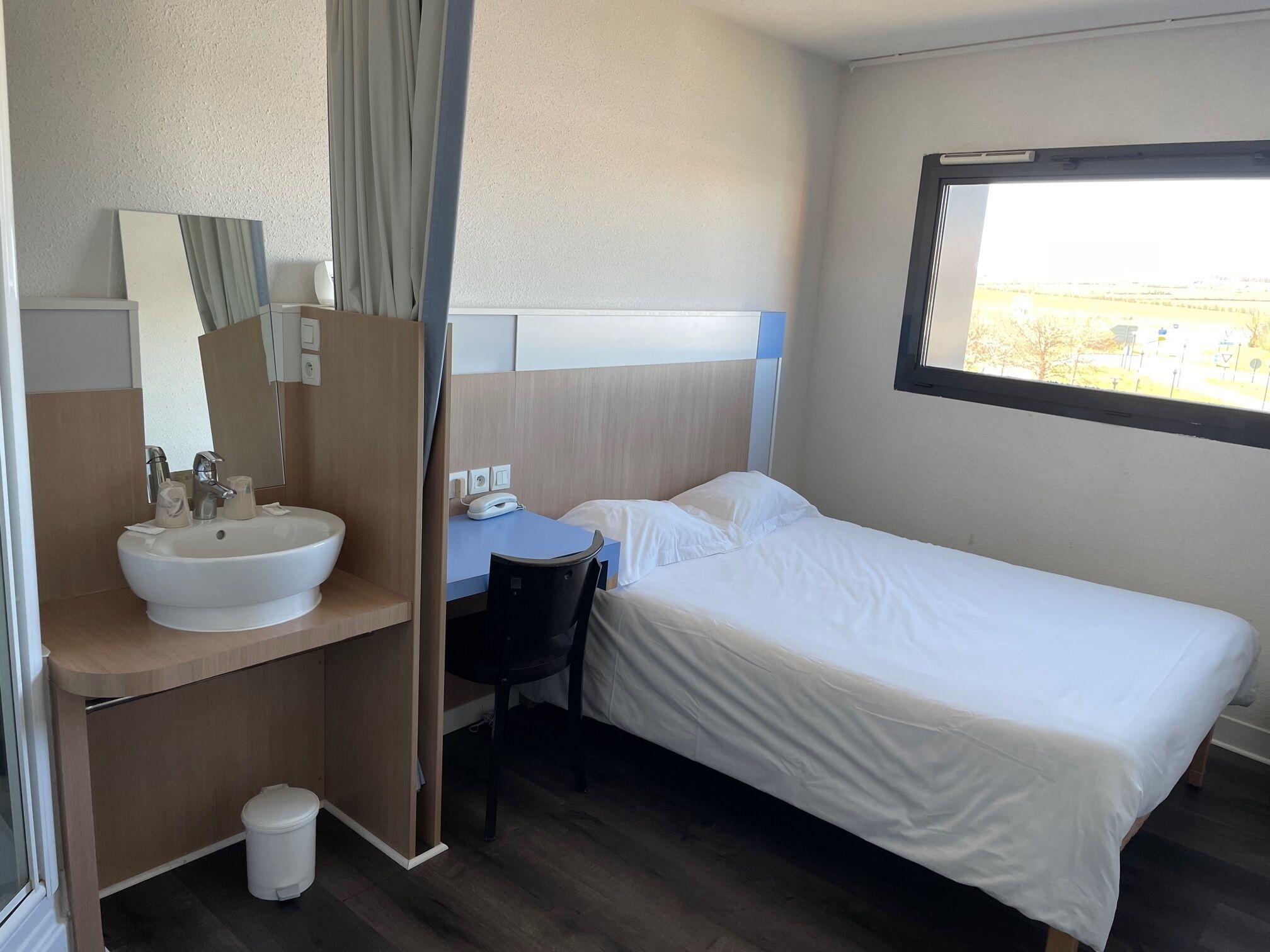 Quarto de hotel com uma cama de solteiro, uma escrivaninha com cadeira, banheiro com pia e espelho, e janela com vista.