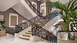 Uma escadaria imponente com grades de metal preto ricamente decoradas, degraus de mármore e um lustre.