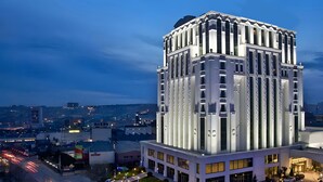 Um hotel alto e moderno com um design vertical marcante e grandes janelas, rodeado por uma paisagem urbana ao entardecer.