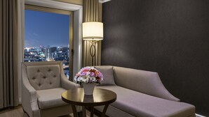 Quarto de hotel moderno com uma grande janela que oferece vista da cidade, um sofá, uma cadeira e uma pequena mesa com luminária e flores.