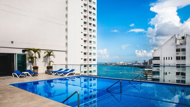 Uma piscina no terraço com azulejos azuis, paredes brancas e vista clara para o oceano e o horizonte da cidade.