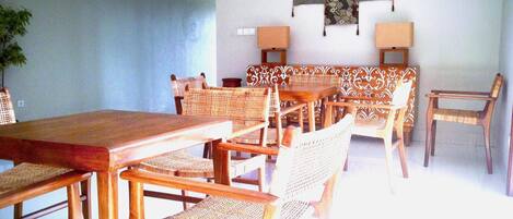 Uma área de refeições com uma mesa de madeira comprida e cadeiras de vime. Há duas luminárias fixadas na parede e uma tapeçaria decorativa sobre a mesa.
