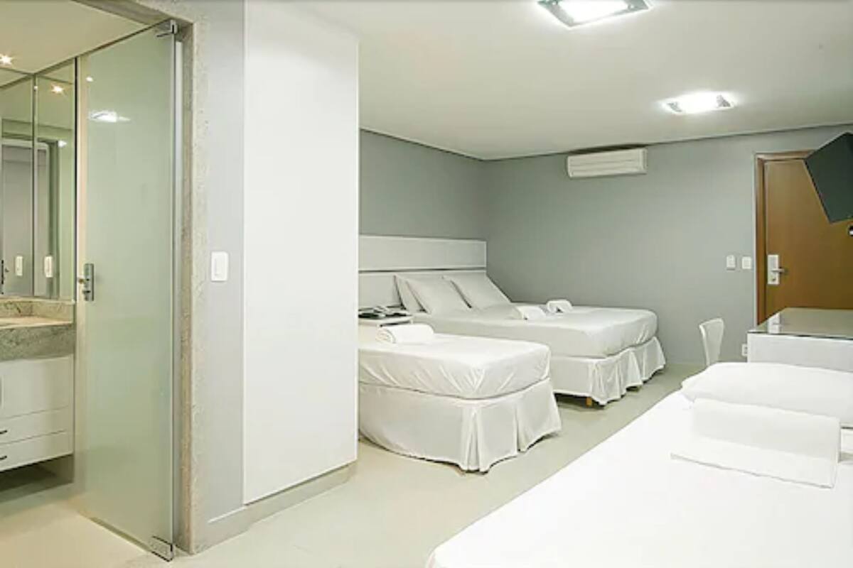 Quarto de hotel com cabine de chuveiro de vidro, duas camas de solteiro, um pequeno espaço para sentar e banheiro com pia e espelho.