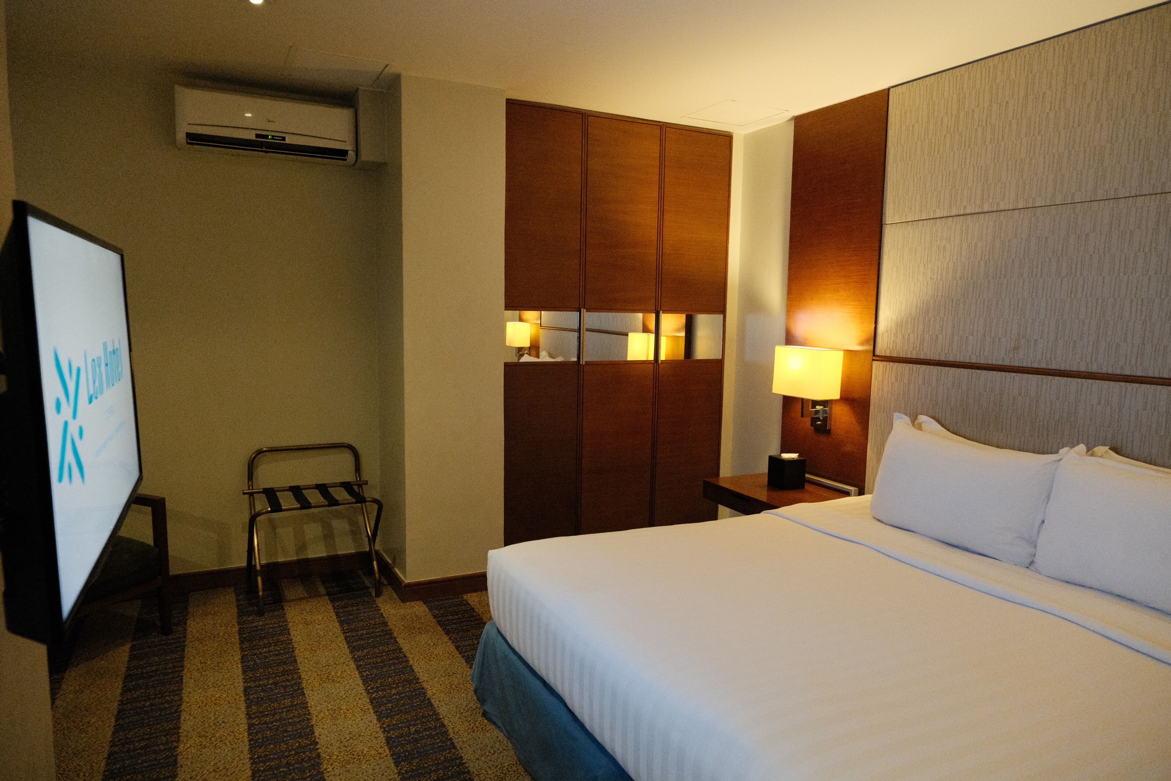 Quarto de hotel com cama, televisão, ar condicionado na parede e um pequeno espaço para sentar.