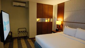 Quarto de hotel com cama, televisão, ar condicionado na parede e um pequeno espaço para sentar.