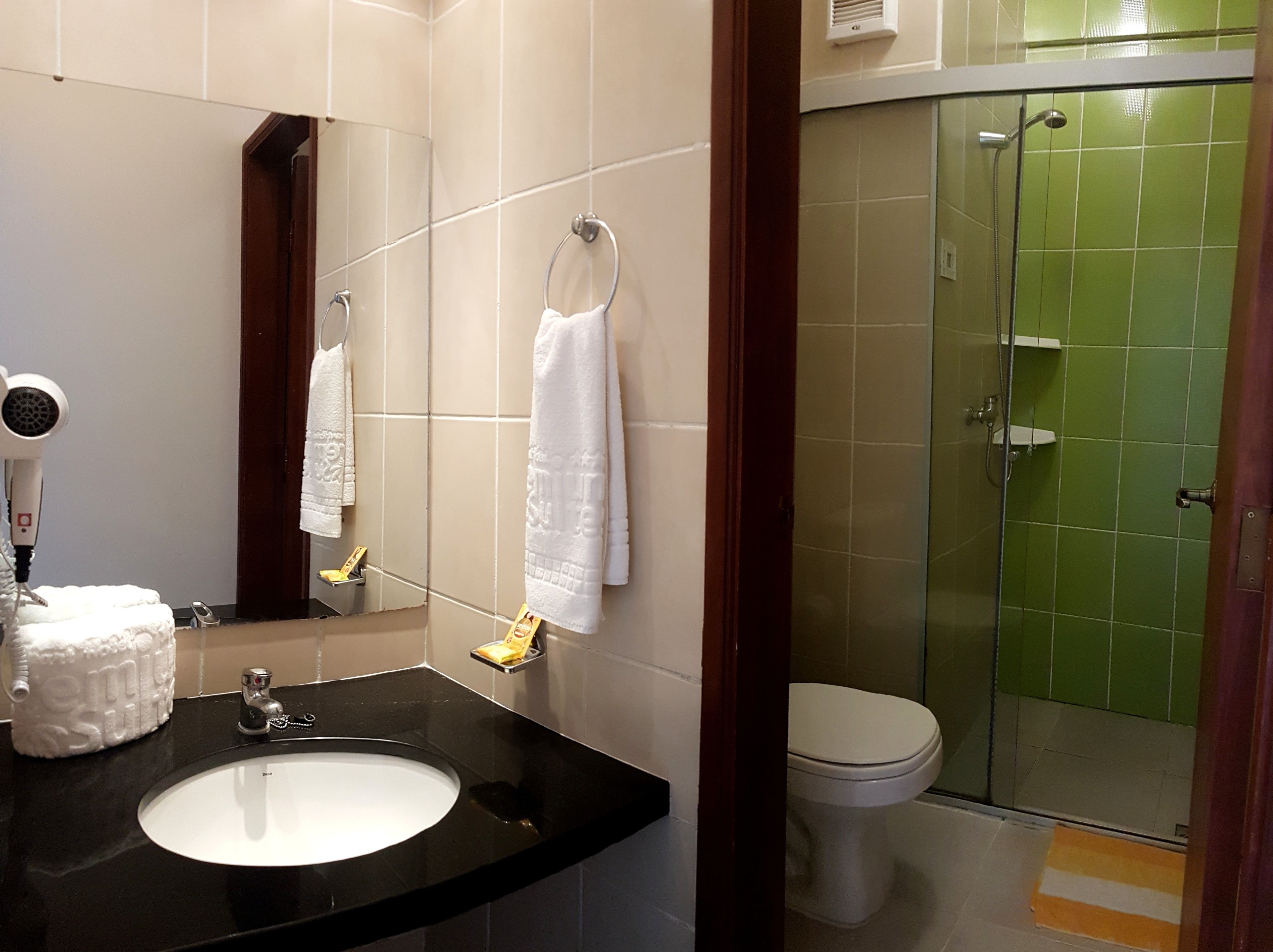 Casa de banho com lavatório preto, toalhas brancas, secador de cabelo e área de duche com azulejos verdes.