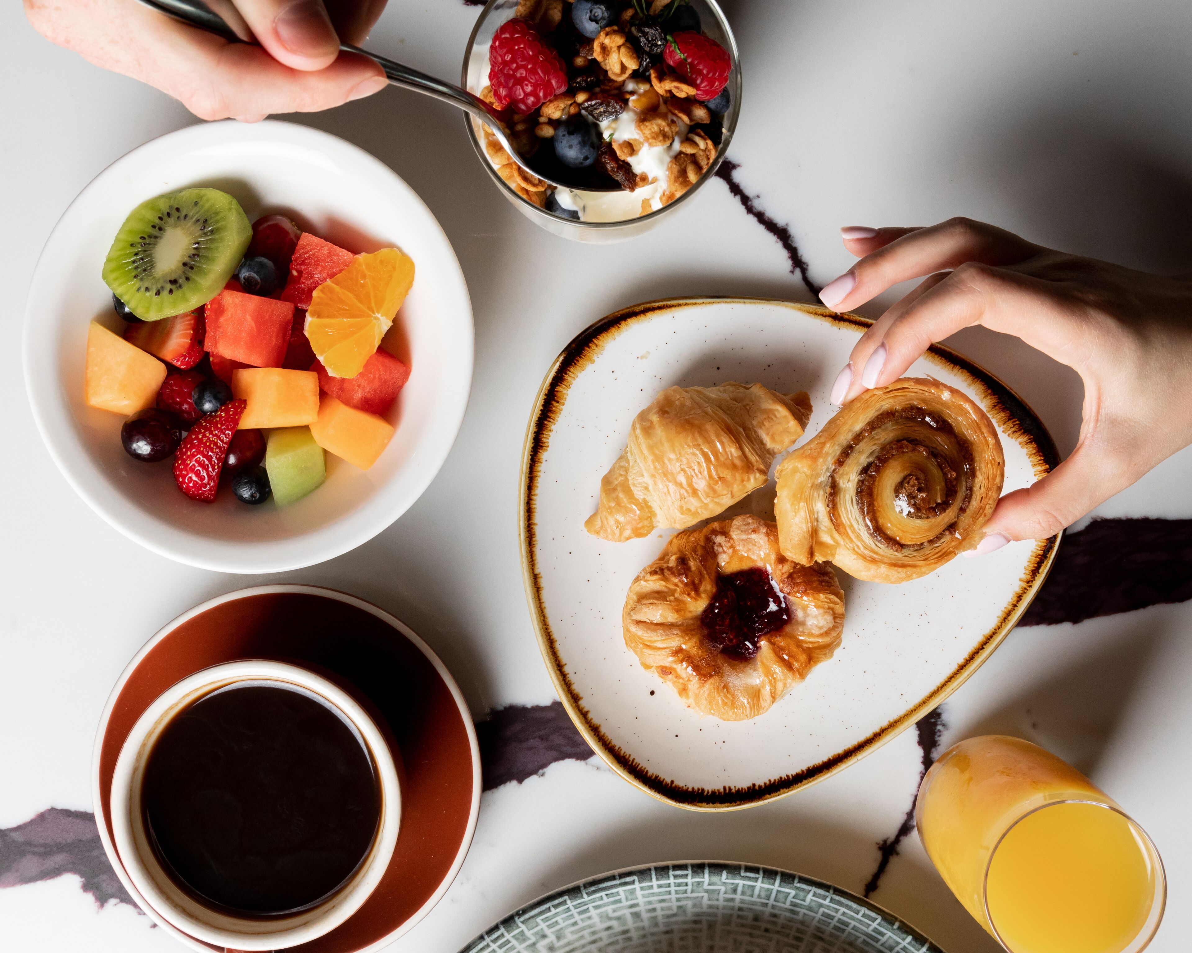 Uma mesa de café da manhã com uma tigela de frutas, croissants, café e suco.