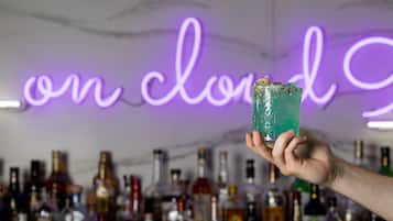 Mão segurando um cocktail verde com cubos de gelo e guarnições, com um letreiro de néon escrito 'on cloud nine'.