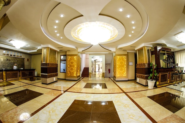 Um saguão de hotel com um candelabro central, balcão de recepção e uma escadaria que leva aos andares superiores.