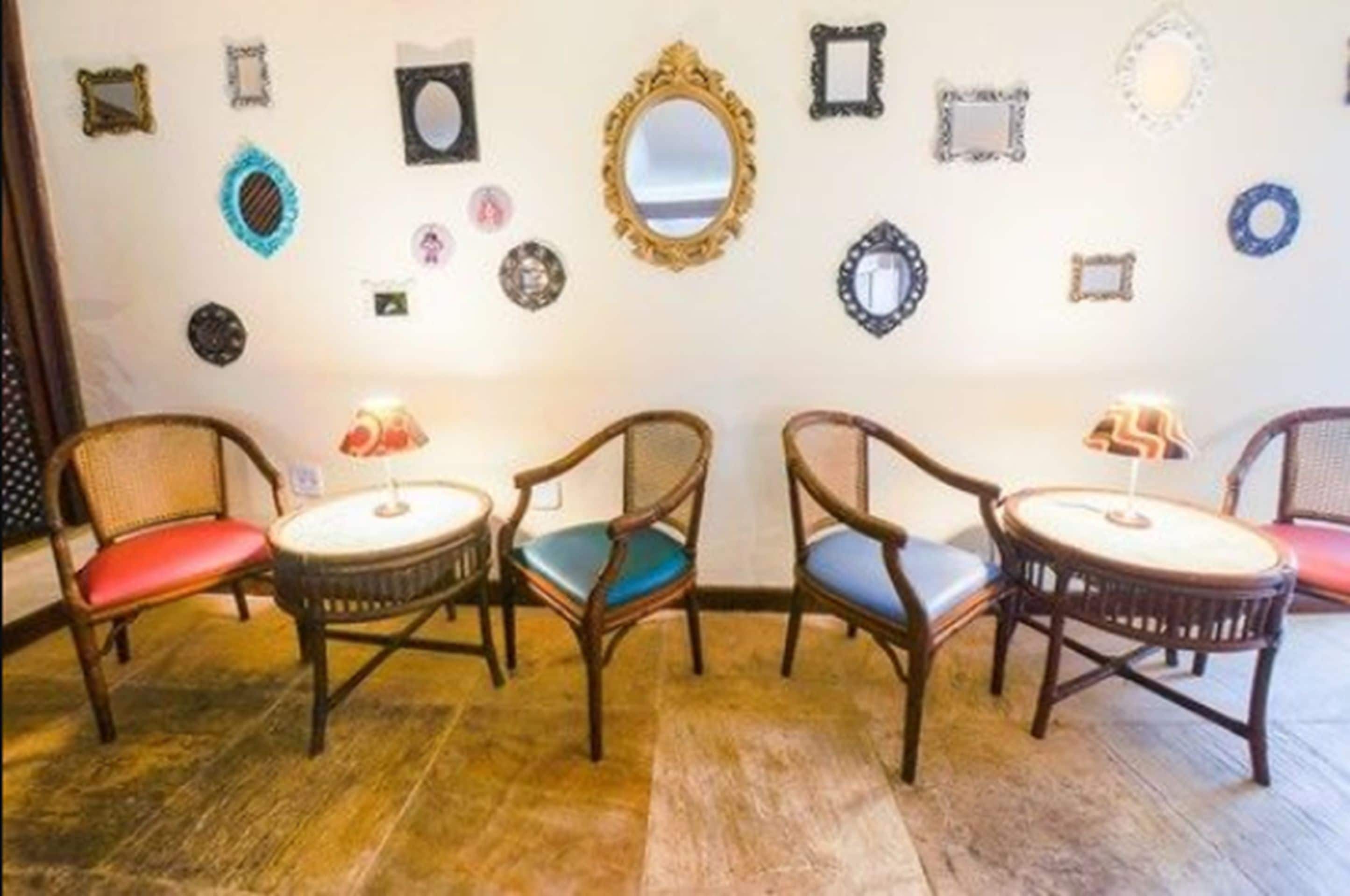 Quarto com quatro cadeiras de vime com almofadas, uma mesa redonda e parede decorada com diversos espelhos emoldurados.