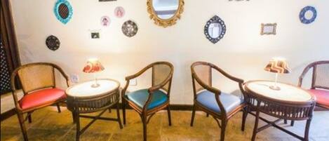 Quarto com quatro cadeiras de vime com almofadas, uma mesa redonda e parede decorada com diversos espelhos emoldurados.