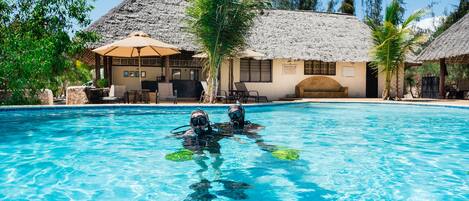 Piscina azul límpida com duas pessoas em equipamento de mergulho, cercada por construções de palha e palmeiras.