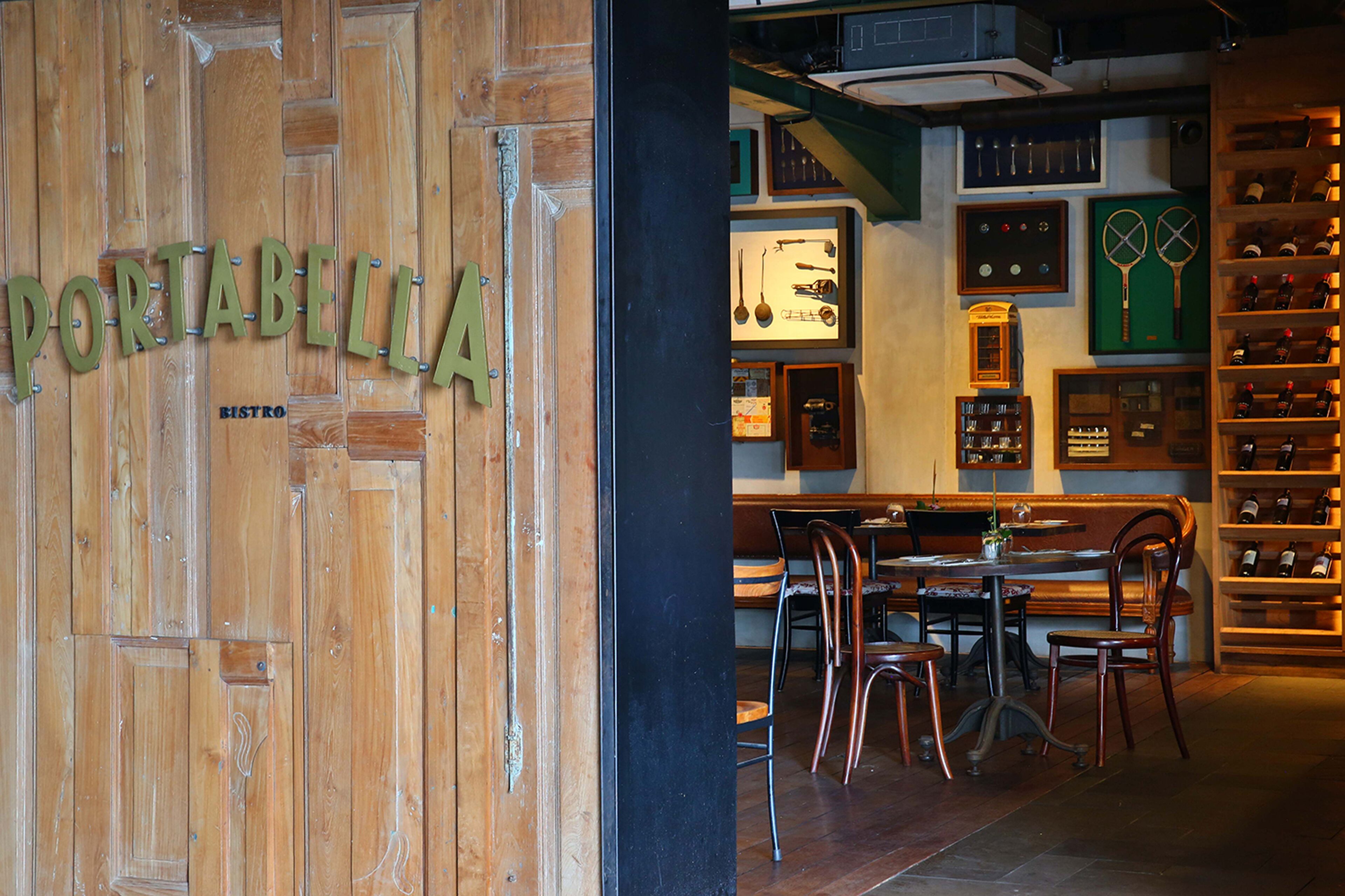 Uma porta de madeira com o nome "PORTABELLA" e uma placa de bistrô. No interior, há mesas e cadeiras, uma exposição de garrafas de vinho e objetos emoldurados na parede.
