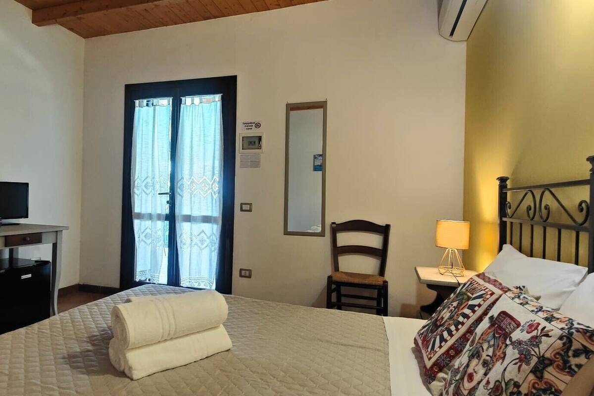 Quarto de hotel com cama, escrivaninha, cadeira, televisão, janela com cortinas e espelho.