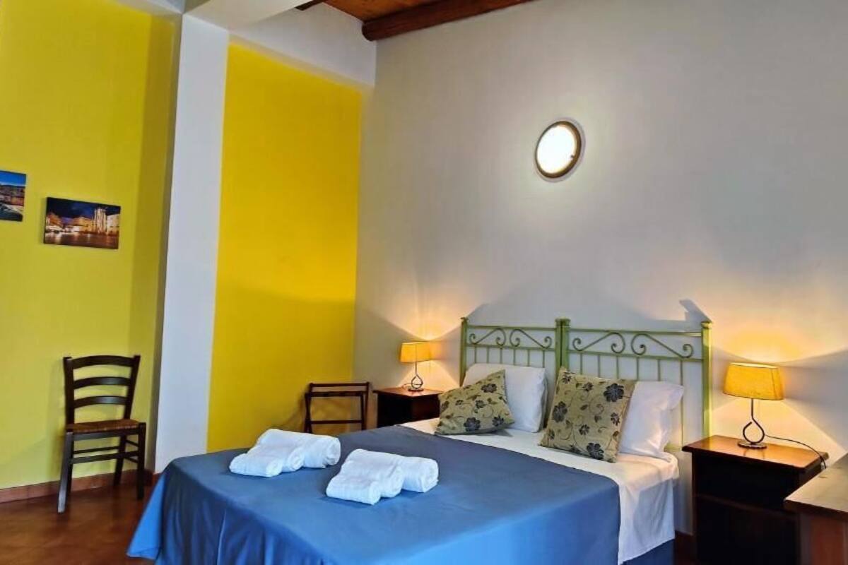 Uma cama bem arrumada com duas almofadas e um cobreleite azul, duas luminárias de cabeceira e uma mesinha com papéis.