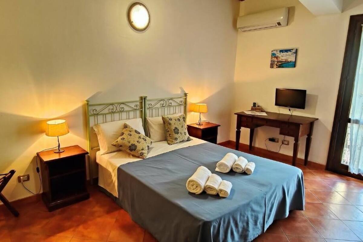 Um quarto com cama, duas luminárias de cabeceira, uma escrivaninha com computador e uma janela.