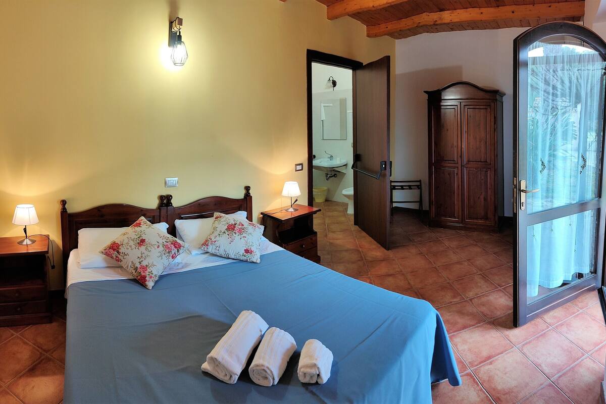 Quarto com cama, duas mesas de cabeceira, um criado-mudo, guarda-roupa de madeira e banheiro visível por uma porta aberta.