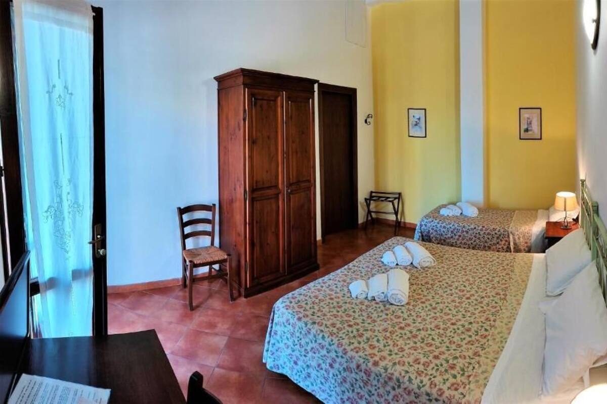 Quarto de hotel com duas camas, um guarda-roupa de madeira, uma cadeira e uma escrivaninha com abajur.