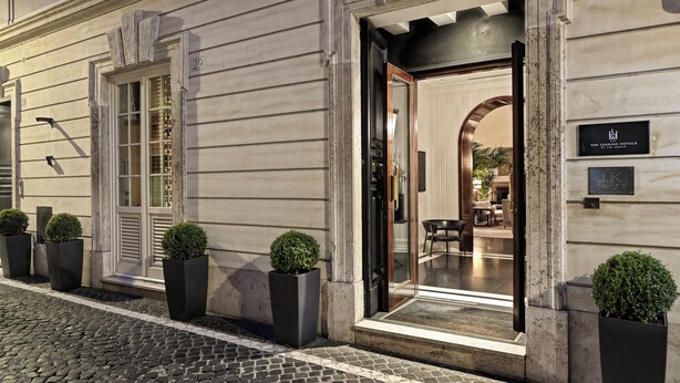Entrada de hotel com rua de paralelepípedos, portas numeradas e interior visível com assentos.