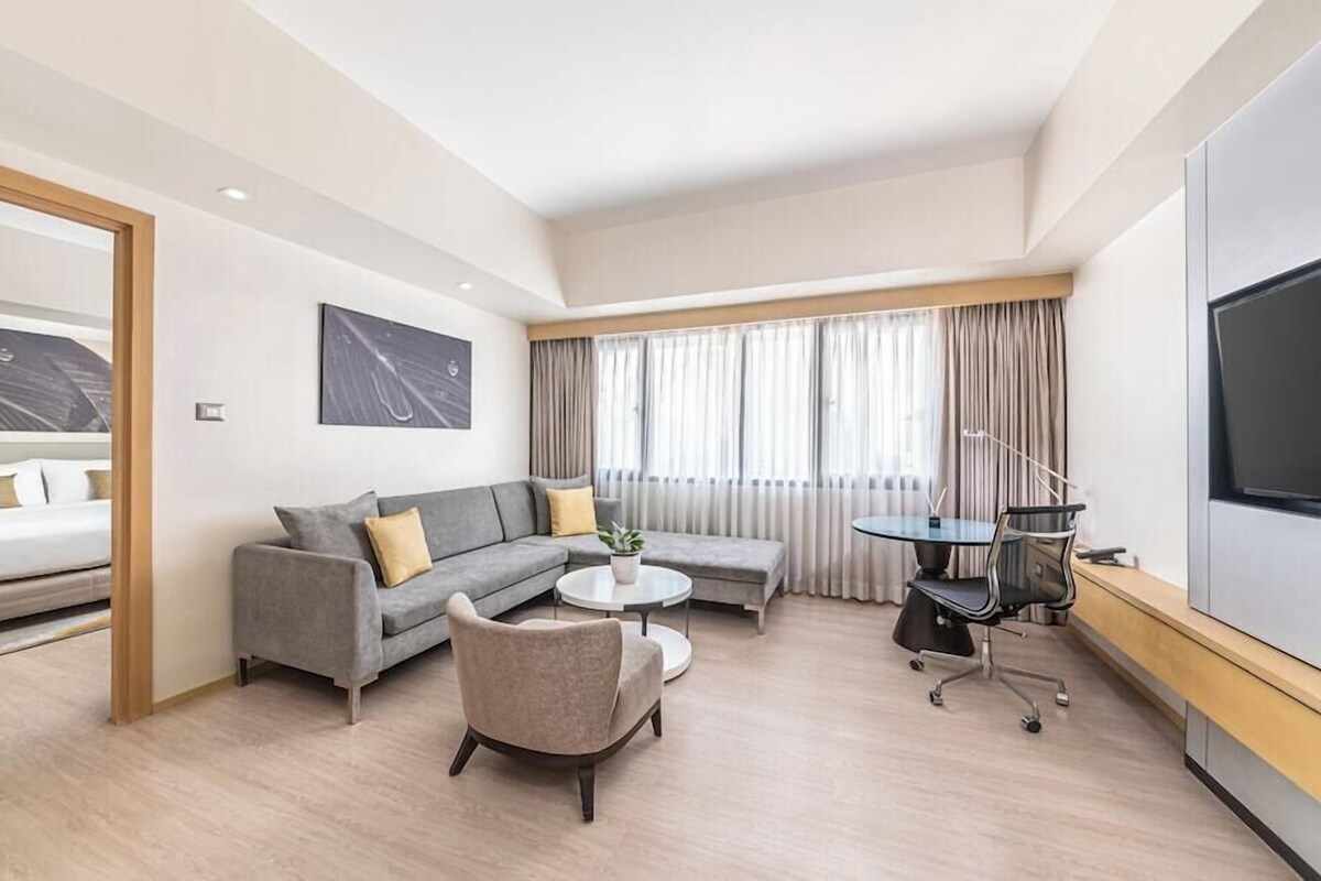 Quarto de hotel moderno com sofá, poltrona, mesa de centro redonda, televisão e uma grande janela com cortinas.