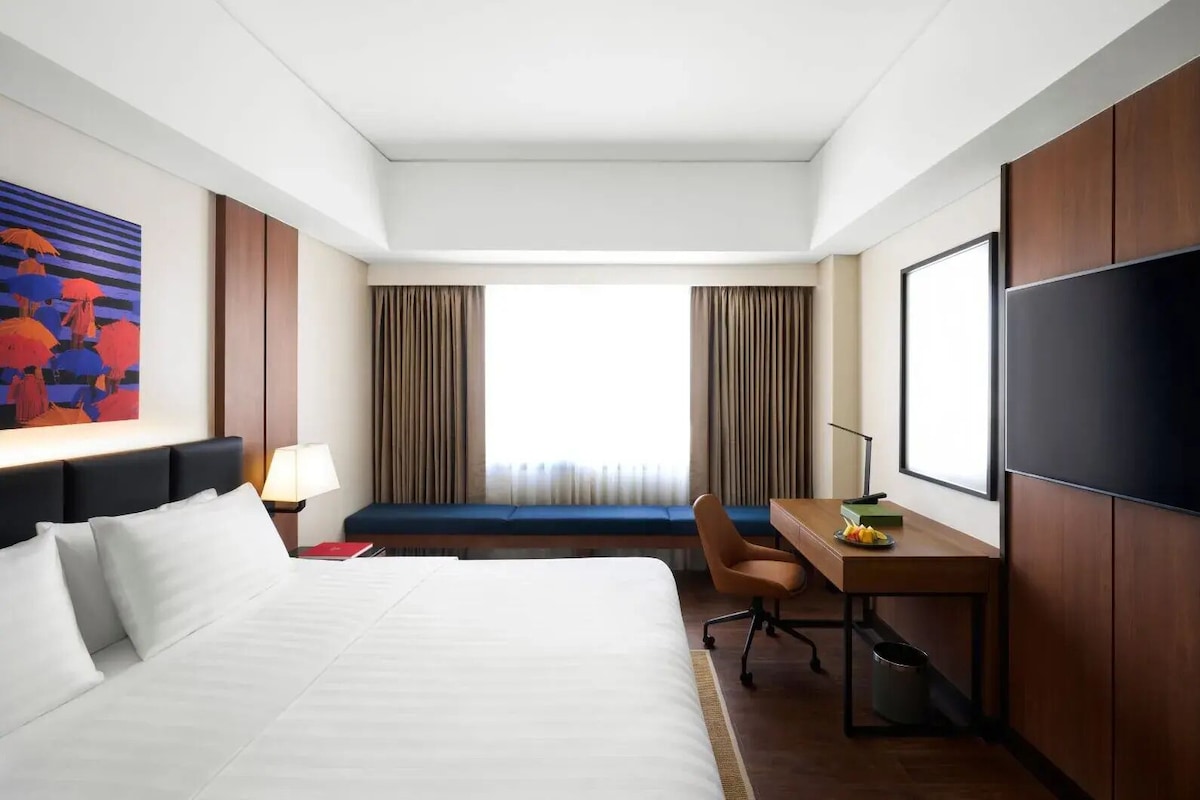 Quarto de hotel com uma cama, escrivaninha, cadeira, televisão e janela com cortinas.
