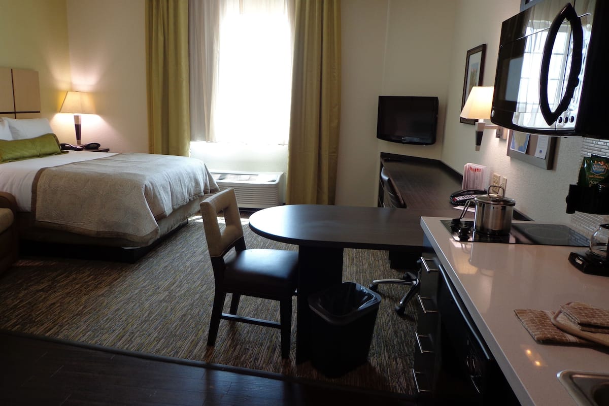 Quarto de hotel com uma cama, uma mesa, uma cadeira, uma TV e uma área de cozinha com fogão.
