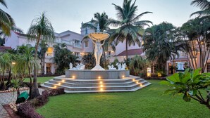 Um jardim bem cuidado com uma fonte central, palmeiras e um edifício com varanda ao fundo.