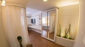 Quarto de hotel com piso de madeira, televisão fixada na parede, porta de vidro deslizante para uma varanda e uma pequena mesa com uma planta.
