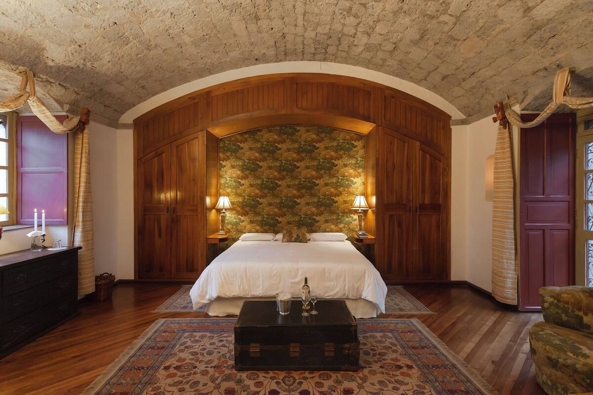 Quarto com uma cama grande, cabeceira de madeira e decoração na parede. Há duas luminárias de cabeceira, um criado-mudo e uma mesinha com uma garrafa e copos.