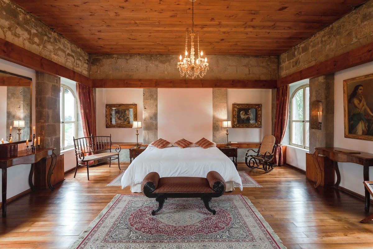 Um quarto espaçoso com uma cama grande, um banco de madeira, um candelabro e uma janela ampla.