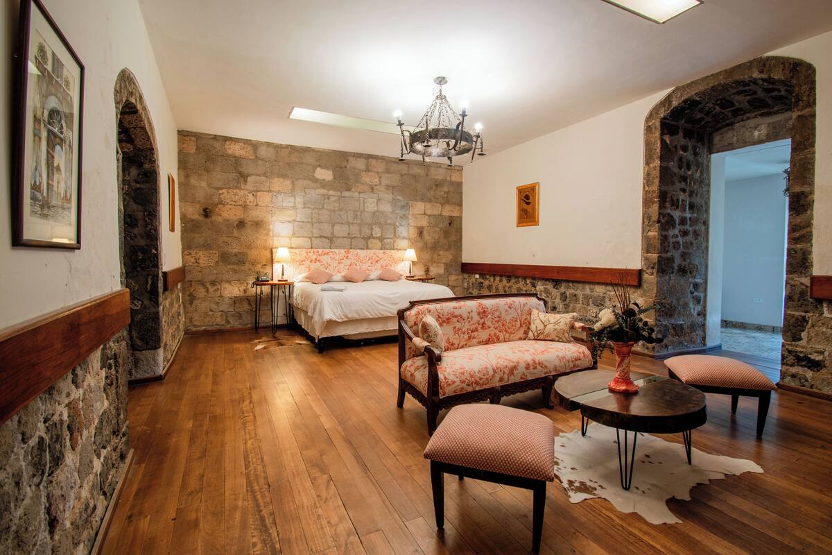 Quarto com parede de pedra, piso de madeira, cama, sofá, poltrona e uma pequena mesa com um vaso.