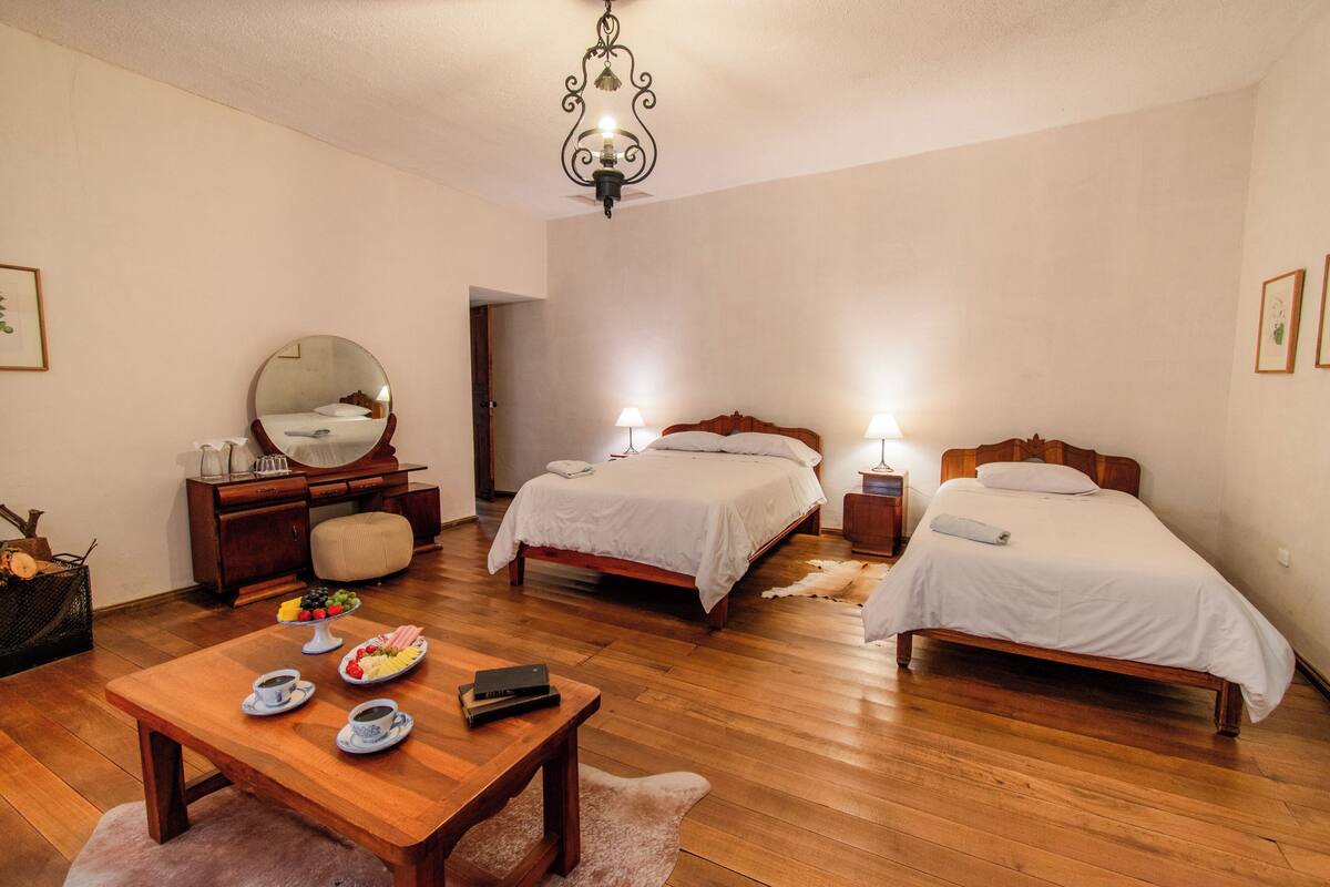 Quarto de hotel com duas camas, uma mesa de madeira com café da manhã, um espelho e um lustre.