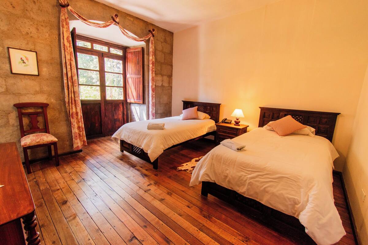 Quarto com duas camas, piso de madeira, uma cadeira, uma mesa e uma janela com cortinas.