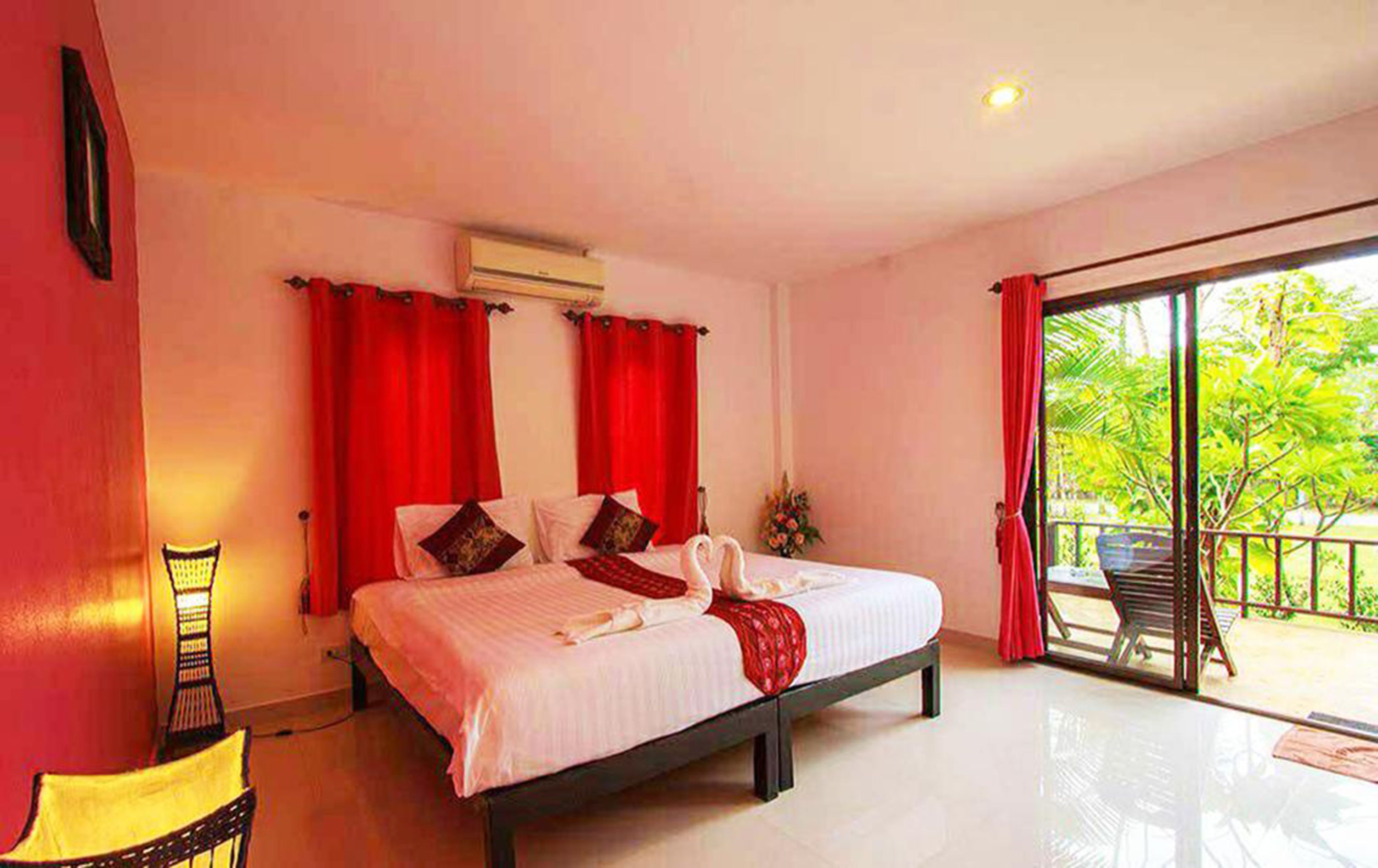 Quarto de hotel com uma cama, uma cadeira, janela com cortinas vermelhas e vista para o verde lá fora.