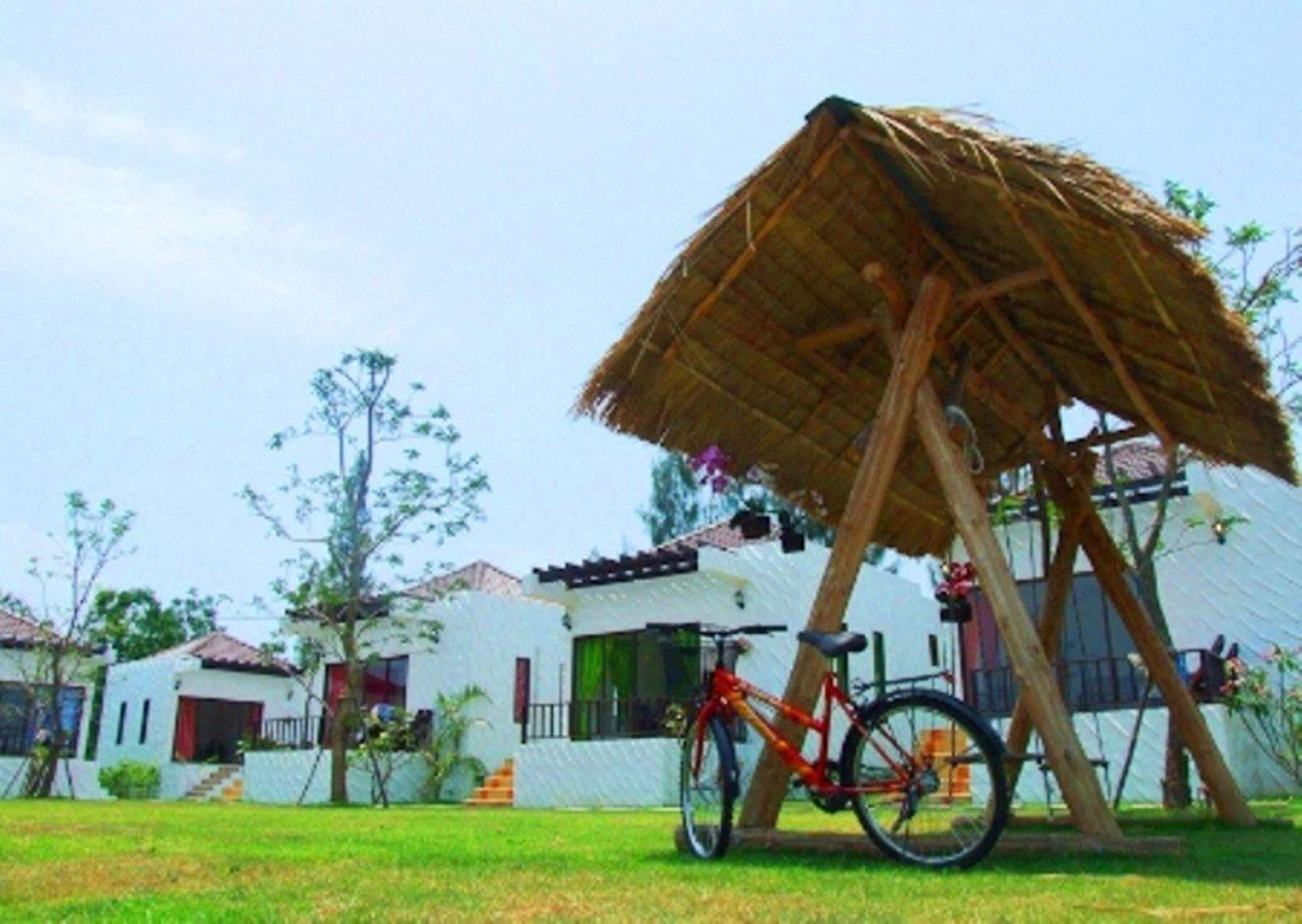 Um edifício branco com estrutura de telhado de palha, uma bicicleta vermelha e um gramado verde.
