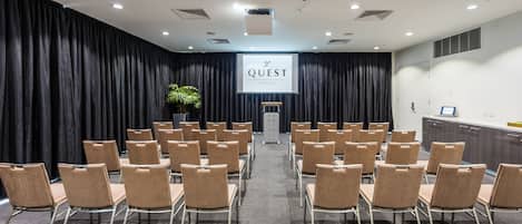 Uma sala de conferências com fileiras de cadeiras, um pódio e uma tela exibindo 'QUEST'.