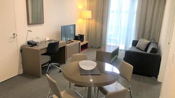 Quarto de hotel com escrivaninha, computador, televisão e área de estar com sofá e poltronas.