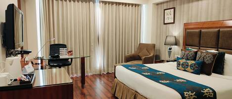Quarto de hotel com cama, escrivaninha com computador, cadeira, abajur e televisão.