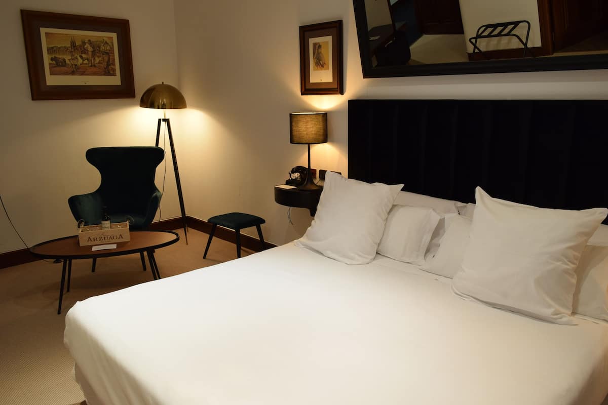 Quarto de hotel com uma cama grande, uma escrivaninha com luminária, uma cadeira e uma pequena mesa com uma garrafa de vinho Arzuaga.
